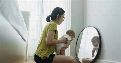장난기 많은 아시아인 어머니는 갓 태어난 아기를 안고 집에서 거울을 보고 앉아 있다 쾌활한 젊은 엄마는 아들과 함께 바닥에서 노는 것을 껴안고 웃고 있다 사진
