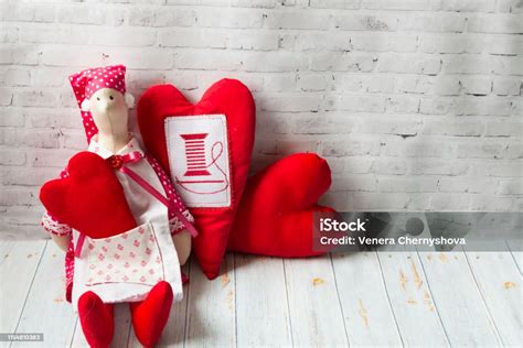 빨간 드레스에 있는 물결 모양의 인형 한 개 흰 앞치마 그리고 빨간 칼 팍에 빨간 섬유 하트가 있는 밝은 배경에 앉아 있습니다 인테리어 인형 텍스트를 위한 장소가 있다