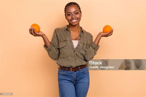 쾌활한 매력적인 사람의 팔은 베이지 색 배경에 고립 된 두 개의 오렌지를 들고 빛나는 미소를 들고 있습니다 땋은 머리에 대한