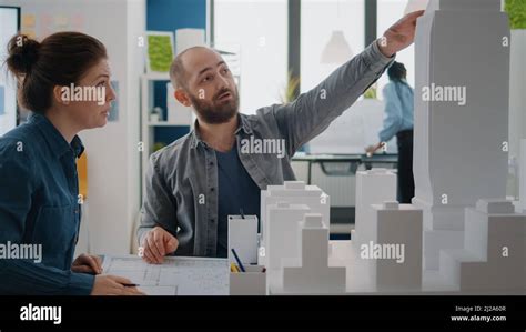 Man And Woman Working Together To Design Blueprints Plan Looking At Building Model On Table