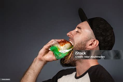 패스트 푸드와 생태의 개념 모자와 수염을 가진 남자가 쓰레기로 채워진 햄버거를 행복하게 먹었습니다 그로테스크하게 확대된 턱 검은 색 배경 복사 공간 남자에 대한 스톡 사진
