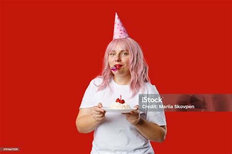 빨간 배경에 케이크를 들고 파이프를 연주하는 생일 파티 모자에 젊은 행복 한 여자 감성적이고 젊은 얼굴 인간의 감정 표정 및