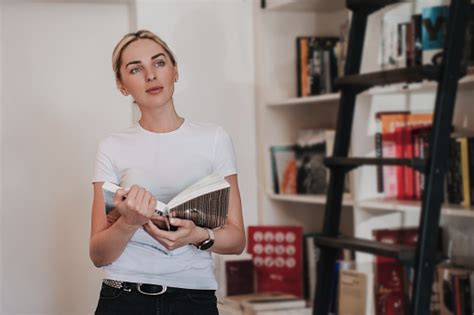 흰색 티셔츠를 입은 잠겨 있는 금발의 똑똑한 젊은 여성이 도서관 계단 앞에 서서 책을 들고 시선을 돌린다 휴식을 취하고 책을 읽는 스칸디나비아 소녀 포니테일 집을 가진 예쁜