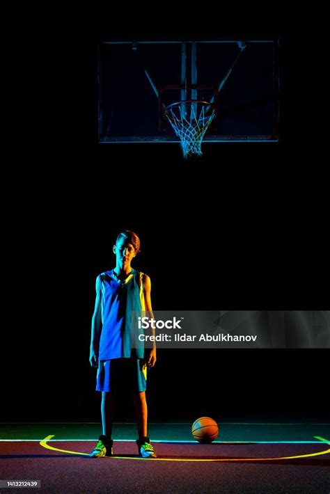 농구 파란색 운동복을 입은 십대가 농구 옆의 놀이터에서 포즈를 취합니다 어두운 배경 멀티 컬러 네온 조명 세로 공간을 복사합니다 스포츠 게임의 개념 놀기에 대한 스톡 사진