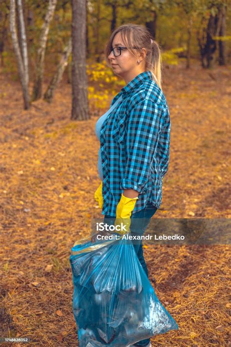 숲에서 쓰레기를 치우고 피곤한 여성 자원 봉사자가 쓰레기 봉투를 손에 들고 서서 슬프게 옆을 바라본다 한 여성이 쓰레기로 인한
