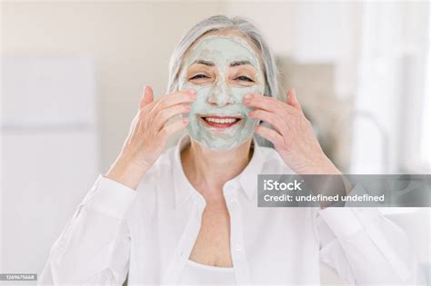 미소 예쁜 선임 회색 머리 여자의 아름다움 초상화를 닫습니다 손으로 그녀의 얼굴에 과일 진흙 영양 얼굴 마스크를 적용 홈 인테리어의 배경에 카메라에 포즈 가정의 방에 대한