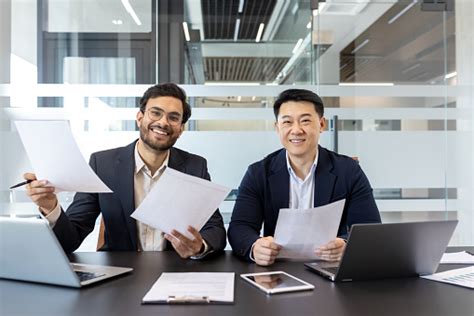 현대적인 사무실에서 노트북과 서류가 있는 책상에 앉아 문서를 들고 웃는 두 명의 사업가가 비즈니스 환경에서 팀워크 협력 및 직업적 성공을 보여줍니다 2명에 대한 스톡 사진