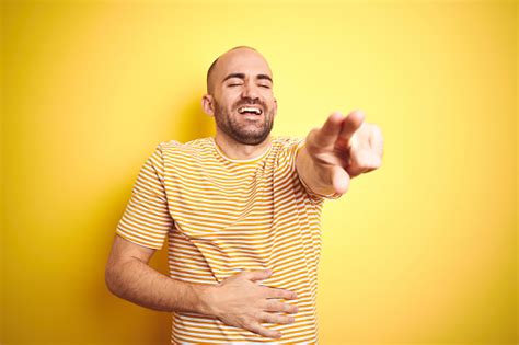 노란 고립 된 배경 위에 캐주얼 스트라이프 티셔츠를 입고 수염을 가진 젊은 대머리 남자는 몸을 손으로 카메라에 손가락을 가리키고 부끄러운 표정을 지었습니다 T 셔츠에 대한