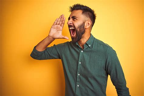 고립 된 흰색 배경 위에 서있는 우아한 셔츠를 입은 젊은 인도 사업가가 입에 손을 대고 큰 소리로 외치고 비명을 지르고 있습니다