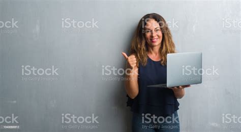 중세 스페인 여자 확인 서명 손가락 우수한 기호를 엄지손가락을 하 고 큰 미소로 행복 노트북을 사용 하 여 회색 그런 지 벽에 서 갈색 머리에 대한 스톡 사진 및 기타