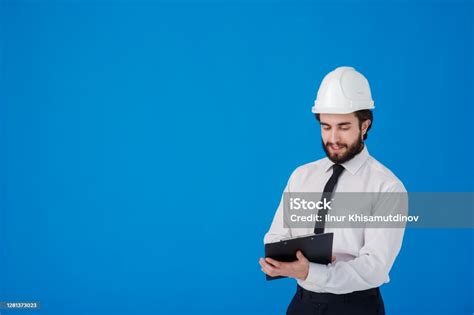 흰색 셔츠와 파란색 배경에 건설 헬멧을 입은 젊은 수염을 기린 남성 건설 노동자 산업 엔지니어건축가는 고립 된 배경에 종이 문서와 클립 보드를 개최 개선에 대한 스톡 사진