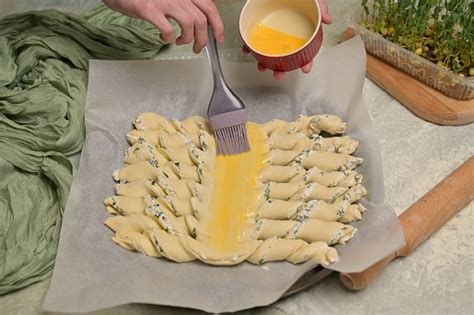 여자의 손이 브러시로 꼬인 반죽 조각에 노른자를 문지른다 계란이 들어간 빨간 작은 그릇 롤링 핀 녹색 수건 부엌 테이블에