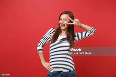 캐주얼 스트라이프 옷을 입고 서 있는 밝은 빨간 벽 배경에 고립 승리 기호를 보여주는 웃는 예쁜 젊은 여자의 초상화 사람들이 진심으 감정 라이프 스타일 개념 복사 공간을