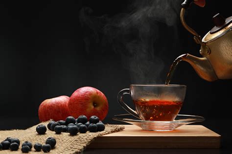 연기와 블루베리가 있는 황금 찻주전자에서 쏟아져 나오는 유리 컵의 뜨거운 차 과일 나무 판에 검은 색 배경으로 사과 건강한 음료
