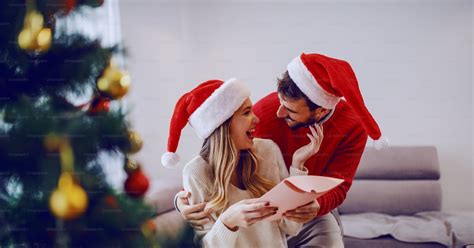 기뻐서 웃고 있는 백인 여자는 거실 소파에 앉아 선물을 들고 남자친구에게 감사를 표한다 여자를 껴안는 남자 둘 다 머리에 산타 모자를 쓰고 있습니다 전경에는 크리스마스