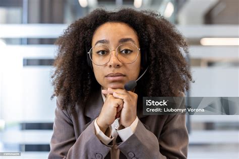 헤드셋과 정장을 착용한 젊은 아프리카계 미국인 여성이 사무실의 책상에 앉아 두 손을 모으고 진지하게 카메라를 바라보는 클로즈업