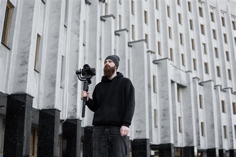 3축 짐벌 안정기에 전문 카메라를 들고 있는 검은 후드를 입은 수염 전문 비디오 그래퍼 전문 영화 카메라로 훌륭한 비디오를 만드는 영화 제작자 촬영 카메라 감독에 대한 스톡