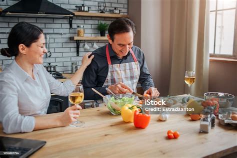 남자는 부엌에 있는 테이블에 앉아서 샐러드 야채를 잘라 그는 모양과 미소 그의 아내 어깨에 손을 잡고 그를 지원 합니다 그녀는 미소 여자 손에 와인의 유리를 있다 가정