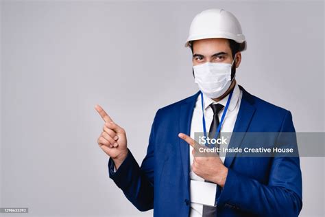 회색 배경 위에 고립 된 복사 공간을 가리키는 코로나 바이러스에 대한 하드 햇과 의료 마스크를 입은 쾌활한 젊은 엔지니어 30 34세에 대한 스톡 사진 및 기타 이미지