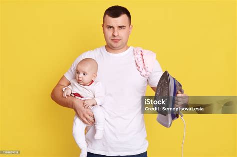 손에 철과 아기를 가진 남자 아버지는 당황 표정으로 카메라를보고 캐주얼 옷을 입고 집안일을하는 유아와 잘 생긴 사람을보고 일을 많이해야합니다 2 5 개월에 대한 스톡 사진