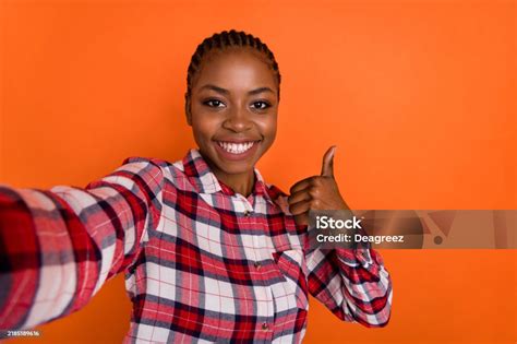 귀여운 자신감 넘치는 숙녀 체크 무늬 셔츠를 입고 엄지 손가락을 보여주는 자체 비디오를 녹화하는 사진 고립 된 밝고 생생한 오렌지 색 배경 땋은 머리에 대한 스톡 사진 및