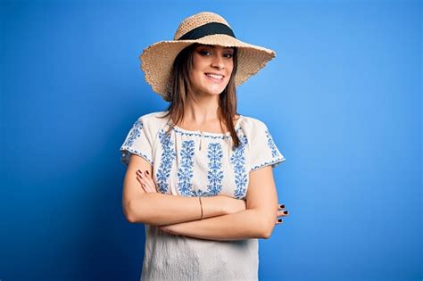젊은 아름 다운 갈색 머리 여자 캐주얼 티셔츠와 여름 모자를 입고 푸른 배경 행복 한 얼굴 카메라를 보고 교차 팔 미소 긍정적