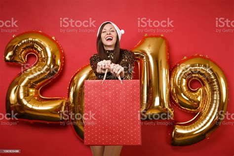 반짝 반짝 드레스에서 메리 산타 여자 크리스마스 모자 밝은 빨간 벽 배경 황금 번호 공기 풍선 스튜디오 초상화에 고립 된 쇼핑백 개최 행복 한 새 해 2019 휴일 파티