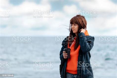 재킷을 입은 젊은 백인 여성의 초상화와 커피 한 잔을 손에 들고 있는 모습이 그려진다 배경에 푸른 바다와 구름하늘 공간을 복사합니다 자유와 여행의 개념 가을에 대한 스톡