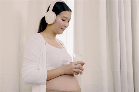 아시아 임산부는 헤드폰을 착용하고 우유 한 잔을 들고 커튼 근처에 서서 임신 중 휴식 자기 관리 건강을 촉진하여 미니멀리즘 가정 환경에서 차분한 환경을 조성합니다 30 34