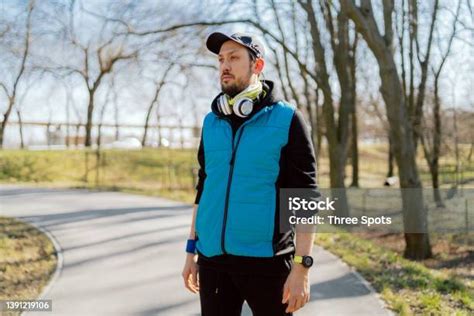 달리기 마라톤을 준비하는 운동 선수의 실루엣 이른 봄 아침에 공공 공원 한가운데에 서있는 모자와 무선 헤드폰이있는 양털 조끼를