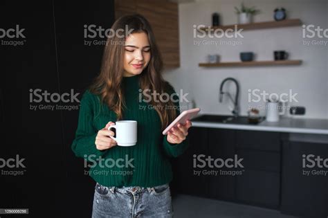 검역 중 부엌에서 젊은 여자 흰색 컵을 잡고 다른 손에 스마트 폰을 보십시오 최신 장치 및 기술 가정 생활에 대한 스톡 사진 및 기타 이미지 Istock