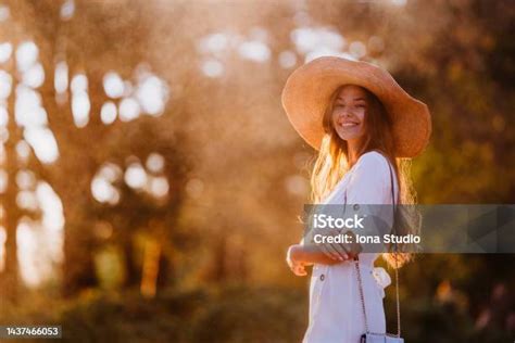 밀짚 모자를 쓴 젊은 호주 여성 여행자 여성 버튼이 달린 흰색 드레스 야생 숲에 서 있는 모자를 만지는 카메라를 보고 있습니다