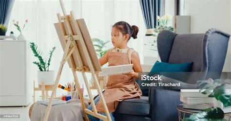 앞치마를 두른 아시아 유아 소녀는 집에서 주말에 추상화를 그리는 데 초점을 맞춘 페인트 브러시를 사용하여 그림 캔버스 앞의 소파 의자에 앉아 있습니다 아이를 위한 창의적인