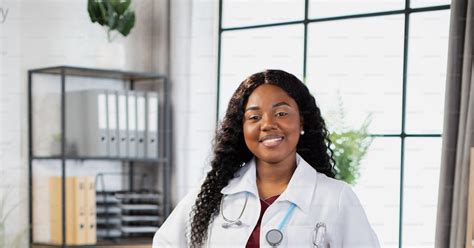 친절한 30세의 예쁜 아프리카계 미국인 여성 의사의 전신 초상화 행복한 미소로 카메라를 향해 포즈를 취하고 그녀의 현대적인 병원 사무실에서 테이블에 기대어 사진