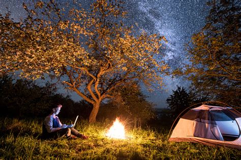 여성 관광 밤에 캠핑에 헤드셋과 함께 그녀의 노트북을 사용 하 여 캠프 파이어와 나무와 아름 다운 밤 하늘 가득 별과은 하 수 아래 텐트 근처에 앉아 여자 관광에 대한 스톡