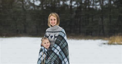 그녀의 아들과 함께 체크 무늬 격자 무늬로 덮인 예쁜 젊은 어머니는 겨울 숲의 배경 위에 서서 크리스마스 화환과 선물 선물로 장식 된 나무 썰매를 당깁니다 사진