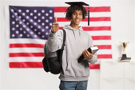 졸업 모자를 쓴 아프리카계 미국인 남학생이 엄지손가락을 치켜들고 미국 국기 앞에서 책을 들고 있다 20 24세에 대한 스톡 사진 및 기타 이미지 Istock