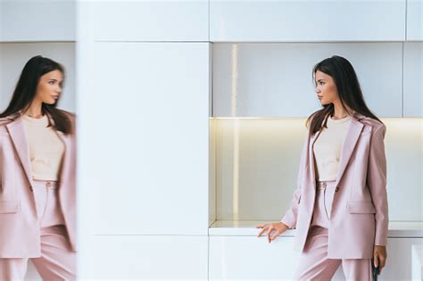 밝은 분홍색 정장을 입은 젊은 목적이 있는 아시아 여성은 미래 지향적인 인테리어에 서서 거울에 자신 있게 반사되어 보입니다 사업가는 집에서 전화를 들고 있습니다 여성