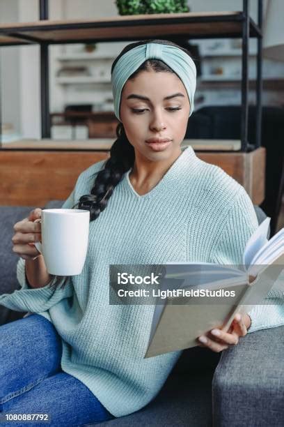 매력적인 혼혈 소녀 청록색 스웨터와 소파에 앉아서 거실에서 차와 독서 책의 컵을 들고 머리 띠 거실에 대한 스톡 사진 및 기타 이미지 Istock
