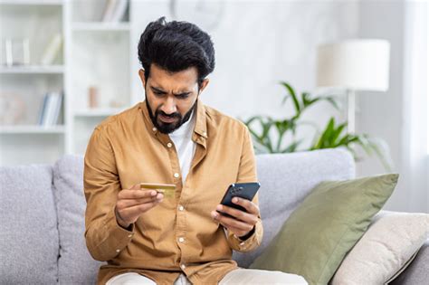 속이고 슬픈 남자는 거실 소파에 앉아서 은행 신용 카드와 전화를 들고 마이너스 계좌 잔고를 들고 거부 된 송금 오류를 받았습니다 20 24세에 대한 스톡 사진 및 기타