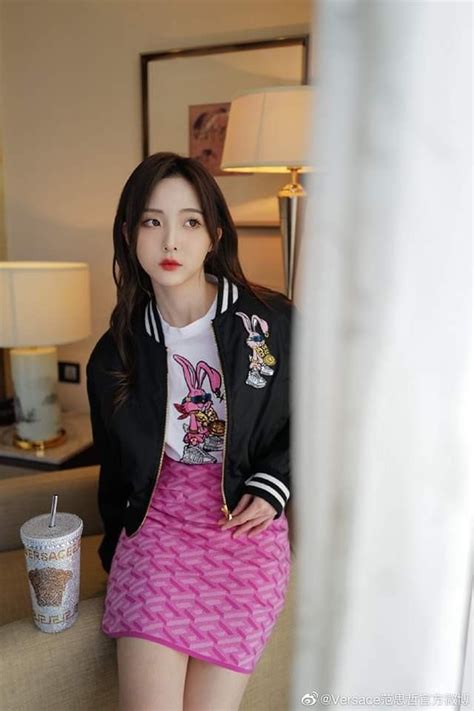 A Woman In A Pink Skirt And Black Jacket Standing Next To A Wall With A Drink