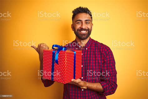 아랍 인도 히스패닉 남자 들고 생일 선물 위에 서 고립 된 노란색 배경 가리키는 고 행복 한 얼굴 미소와 함께 측면에 엄지 손가락으로 표시 가리키기에 대한 스톡 사진 및