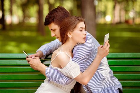 Gadget Addicted Couple Using Mobile Phones While Embracing On Bench At