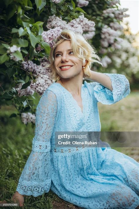 Blonde Woman In A Blue Dress Sitting Near A Flowering Bush High Res