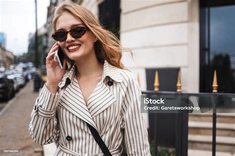 줄무늬 트렌치코트를 입은 젊은 예쁜 웃는 여자가 아늑한 도시 거리를 걷는 핸드폰에 행복하게 이야기합니다 16 17 살에 대한 스톡 사진 및 기타 이미지 Istock