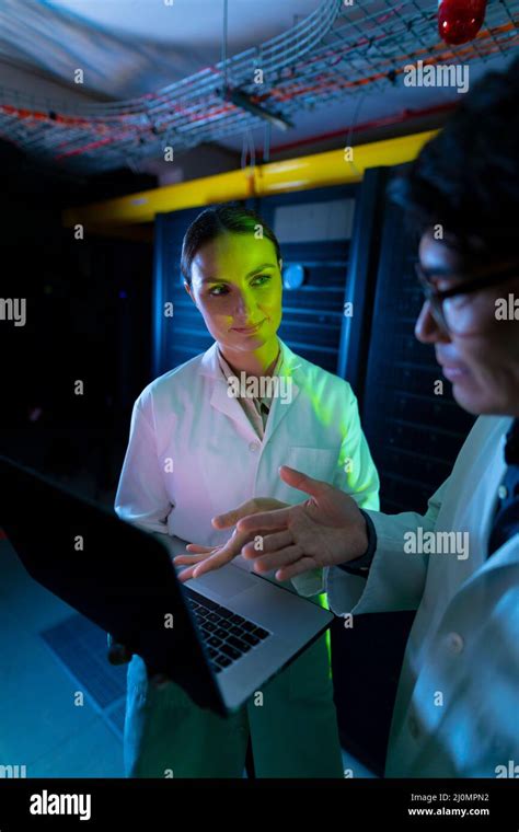 Diverse Male And Female Engineers Wearing Aprons Discussing Over Laptop In Computer Server Room