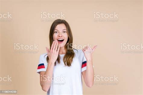 파스텔 베이지 색 배경에 서있는 젊은 아름답고 흥분한 여성이 손바닥을 얼굴 근처에 들고 손가락을 가리키고 있습니다 가리키기에 대한 스톡 사진 및 기타 이미지 Istock