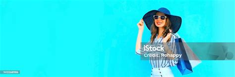 하얀 줄무늬 셔츠를 입고 멀리 바라보는 쇼핑백을 가진 아름다운 미소 젊은 여성의 초상화 파란색 배경에 여름 검은 둥근 밀짚 모자 광고 텍스트를위한 배너 빈 복사 공간 25