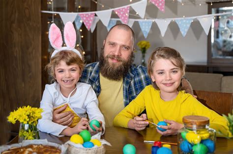 부활절 가족 전통 아버지와 토끼 귀를 가진 두 명의 백인 행복한 아이들은 집 테이블에 함께 앉아 휴일을 위해 페인트로 계란을 염색하고 장식합니다 아이들은 아늑하게 포옹하고