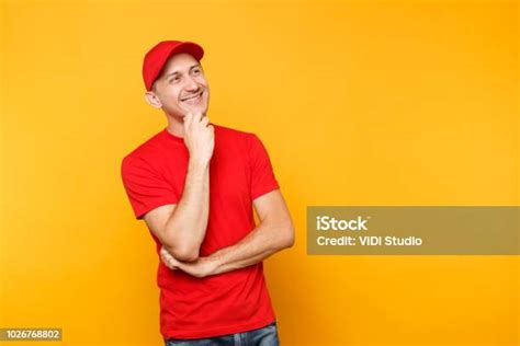 빨간색 유니폼 노란색 오렌지 배경에 고립 된 배달 남자 전문 모자에서 남성 직원 미소 T셔츠 택배 또는 딜러로 일하고 올려 손 형은 턱에 서비스 개념입니다 공간에 복사